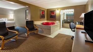 Quarto de hotel com cama, duas poltronas, escrivaninha, televisão, banheira vermelha e espelho.