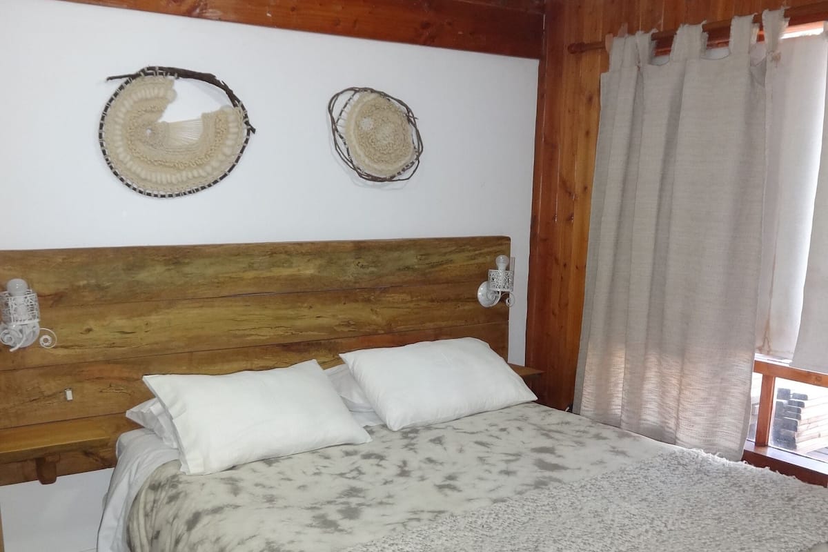 Quarto com cabeceira de madeira, cama com roupa de cama branca, duas decorações de parede e uma janela com cortinas.