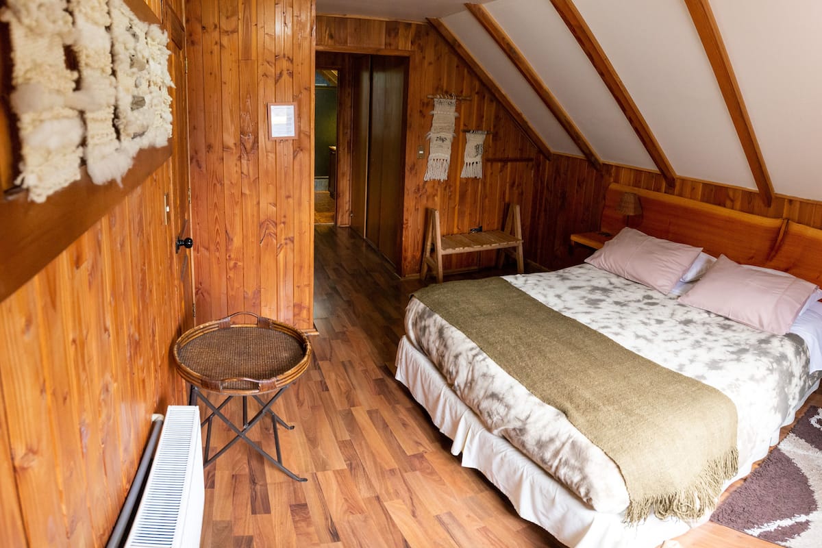 Um quarto de sótão aconchegante com paredes de madeira, uma cama com colcha estampada, uma cadeira e um radiador.