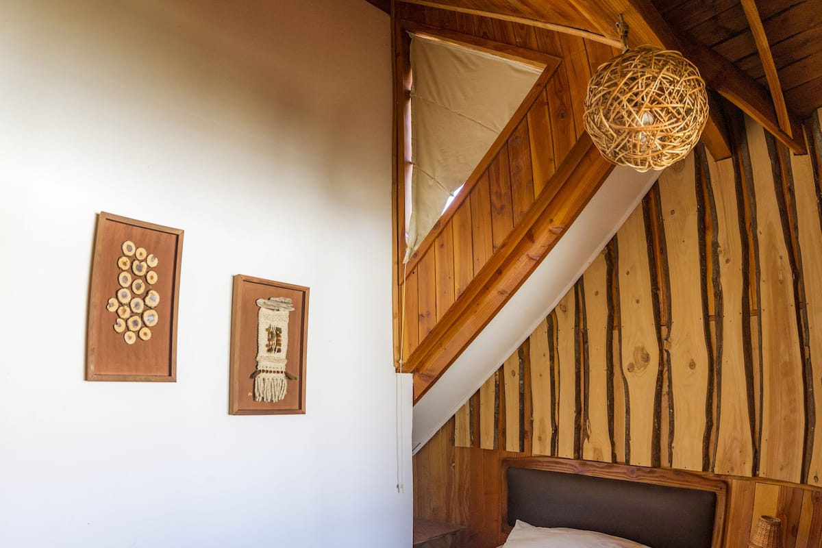 Quarto com teto de madeira, cama com roupa de cama branca, mesa de cabeceira pequena e luminária fixada na parede.