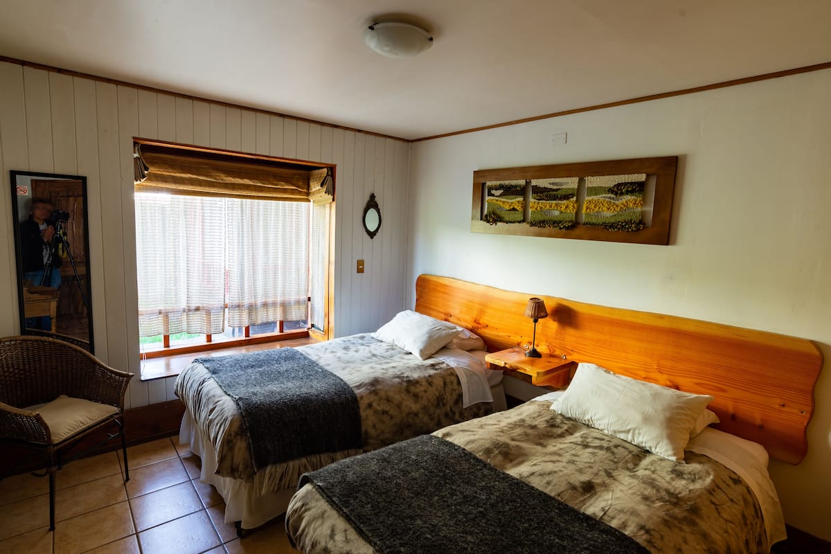 Quarto com duas camas, cabeceira de madeira, janela com cortinas, uma cadeira de vime e um quadro na parede.