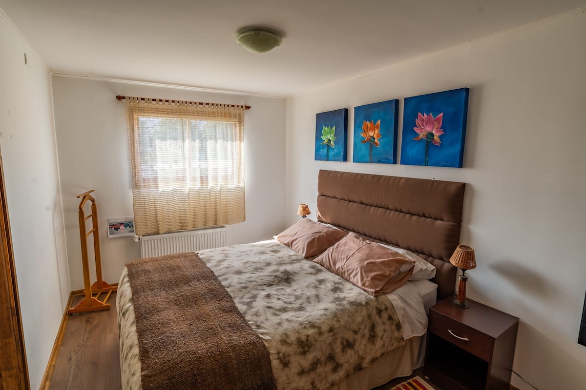 Quarto com cama, mesa de cabeceira, luminária, janela com cortinas e três quadros pendurados na parede.