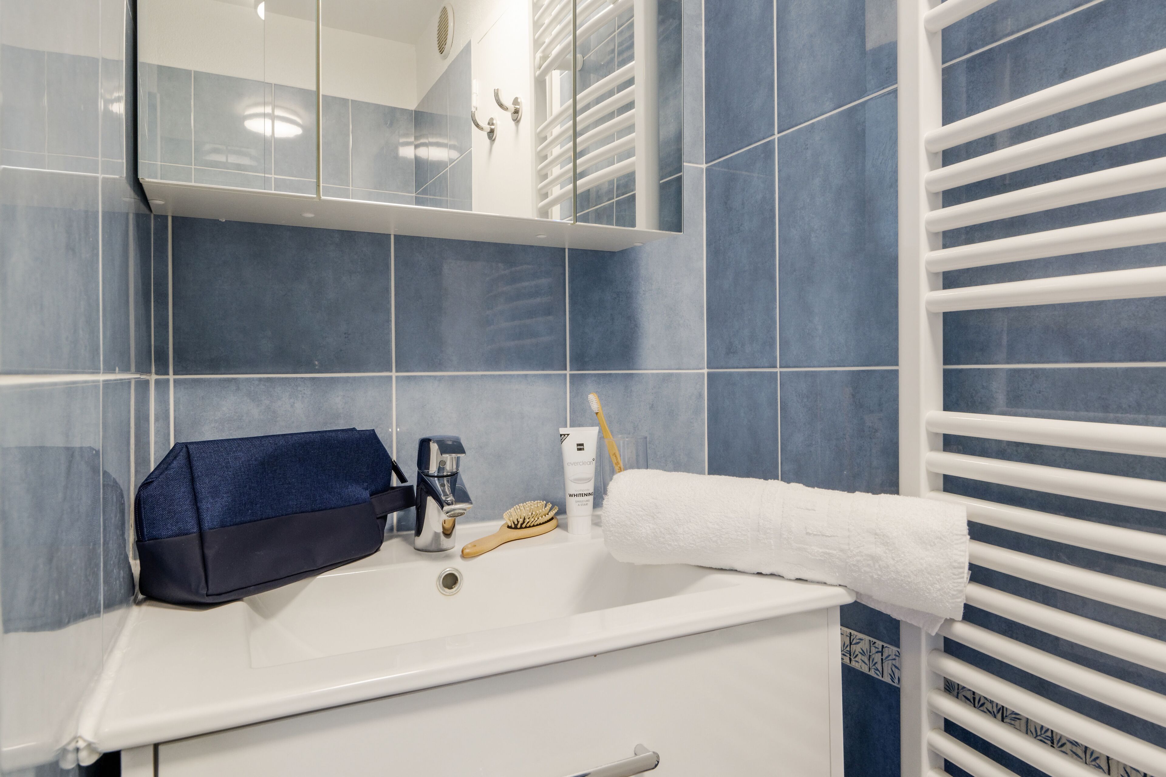 Casa de banho com uma pia branca, azulejos azuis, uma toalha, uma escova de dentes e uma mala azul.