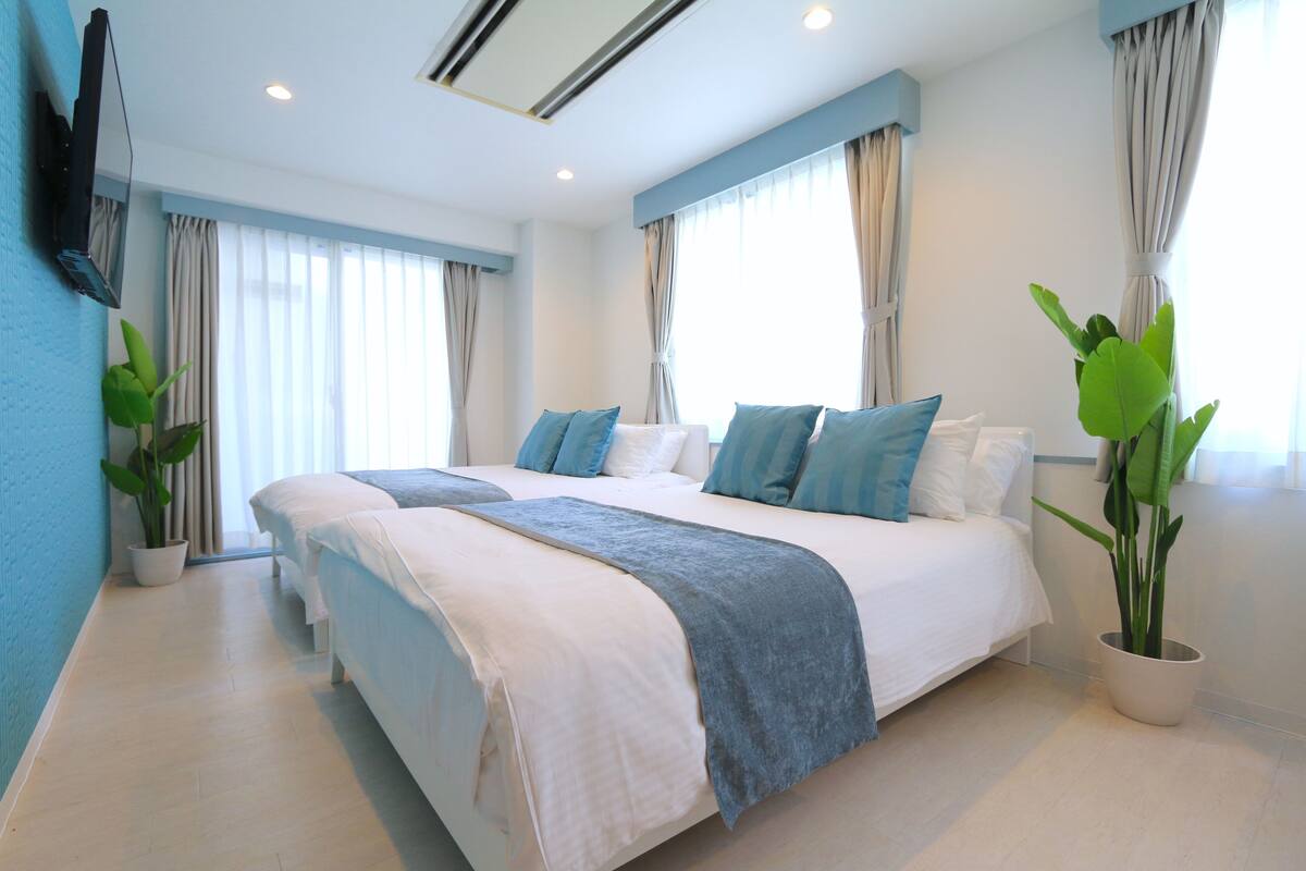 Uma cama de casal com roupa de cama branca e azul, duas plantas em vasos e uma televisão de tela plana na parede.