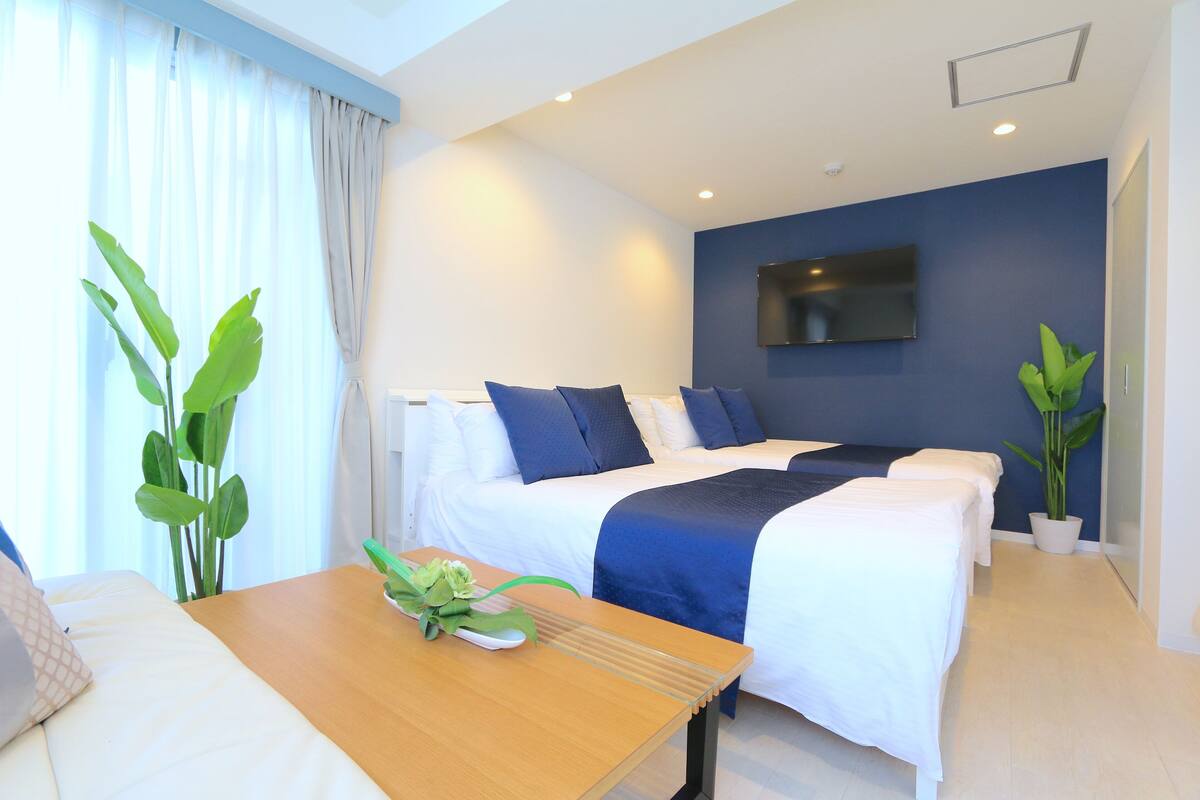 Quarto de hotel moderno com uma cama grande, uma cama menor, uma mesa de madeira com uma planta e uma TV fixada na parede.