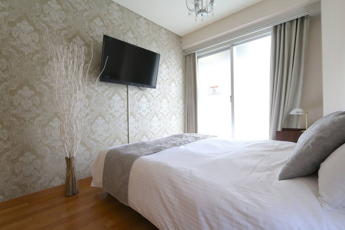 Quarto com cama, televisão de tela plana na parede, janela com cortinas e um vaso com galhos.