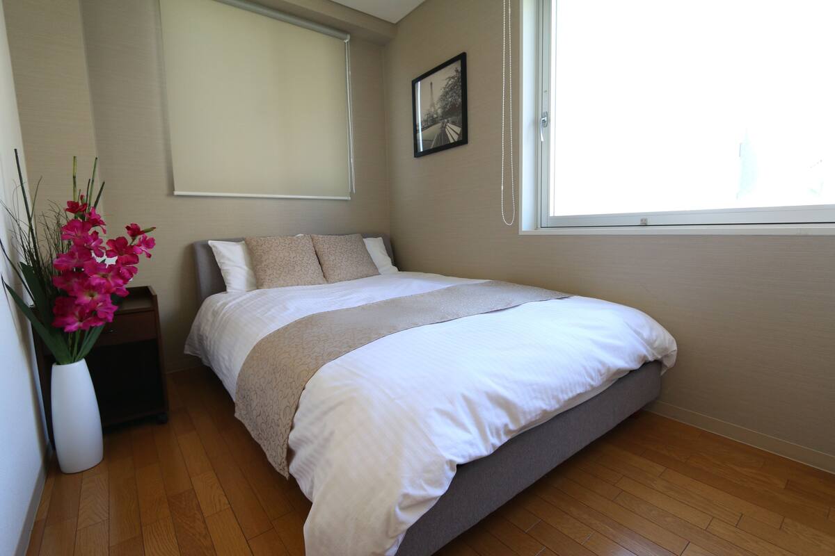 Quarto com cama, mesa de cabeceira, vaso com flores, janela e quadro na parede.