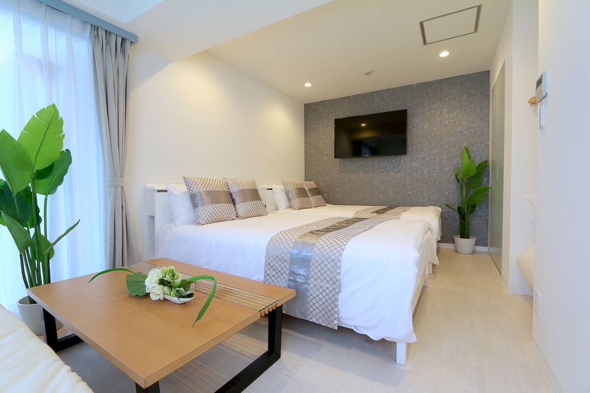 Quarto de hotel com cama, mesas de cabeceira, televisão presa na parede e plantas em vasos.