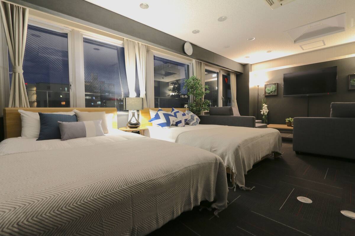 Quarto de hotel com duas camas, um sofá, uma televisão e janelas amplas.