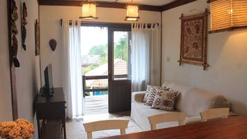 Sala de estar com sofá, televisão em móvel, mesa de madeira e vista para o exterior através de uma porta de vidro.