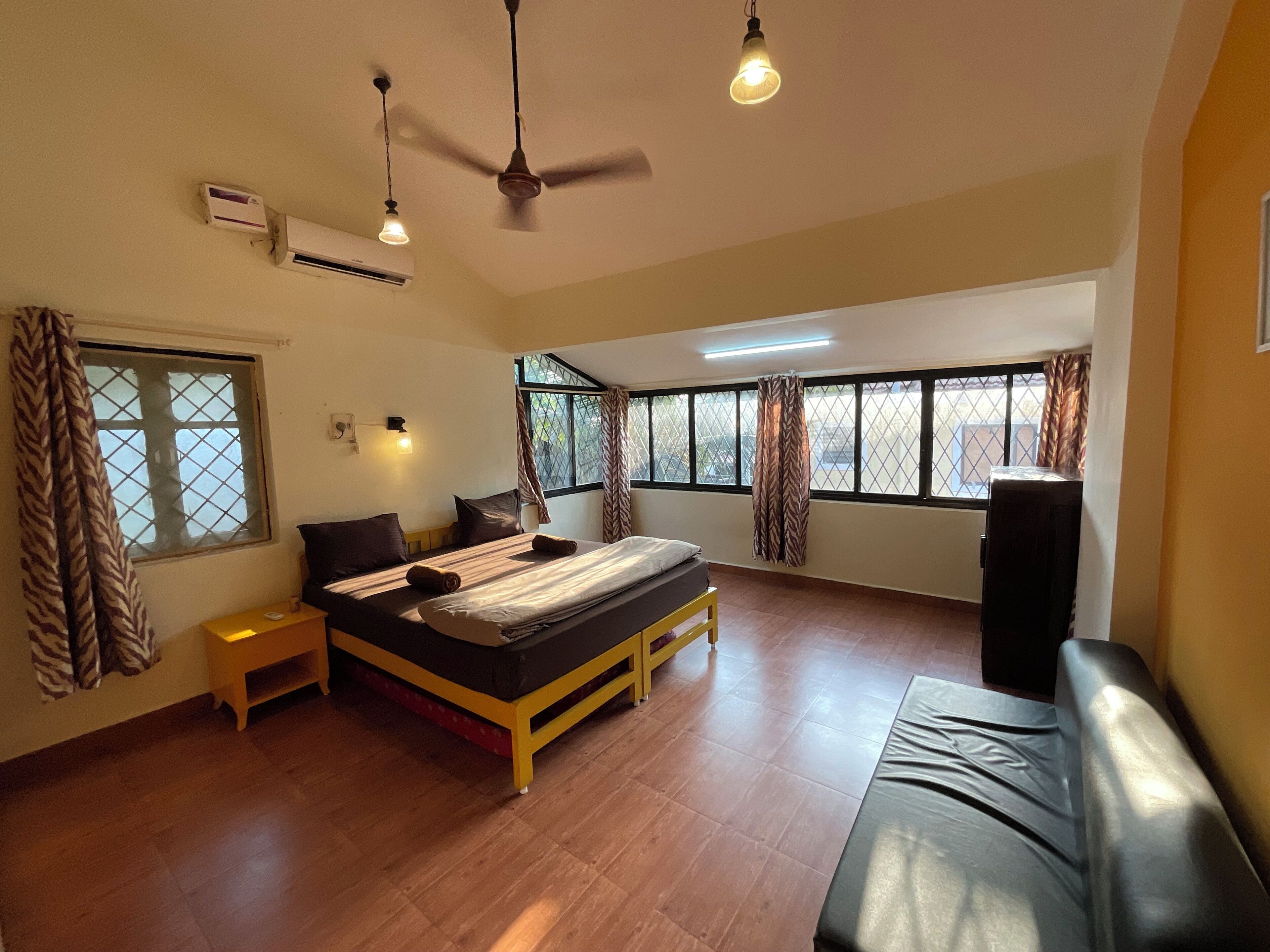 Quarto com cama, mesa de cabeceira, banqueta, ventilador de teto, ar condicionado e janelas amplas com cortinas.