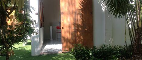 Uma pequena área externa cercada, com uma porta de madeira, uma parede branca e uma pequena mesa com uma cadeira.