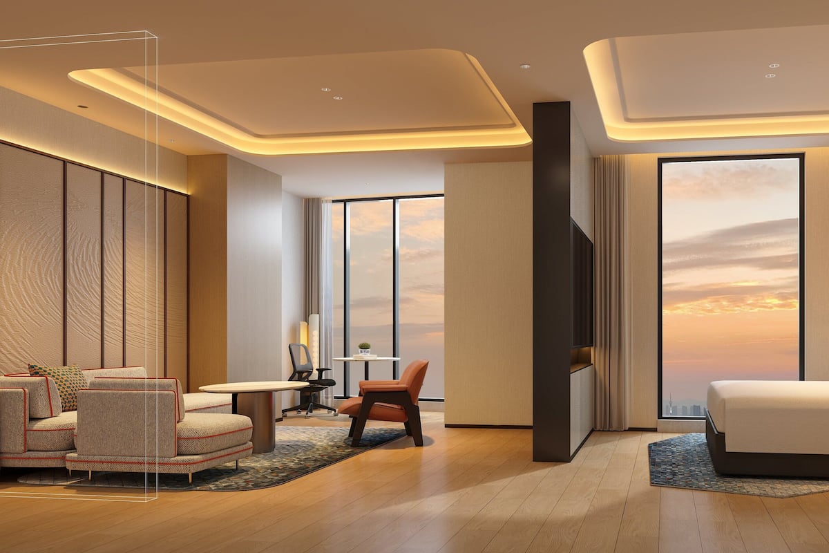 Quarto de hotel moderno com cama grande, área de estar com sofá e poltronas, pequena mesa e uma janela ampla com vista para a cidade.