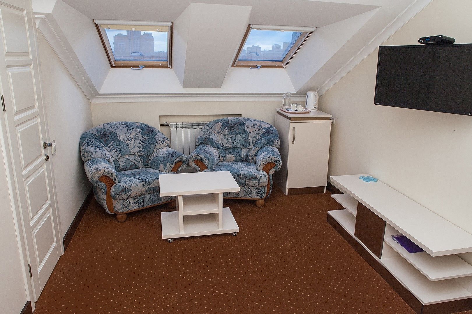 Quarto compacto com duas poltronas estampadas, uma mesinha branca, um armário e uma TV fixada na parede.
