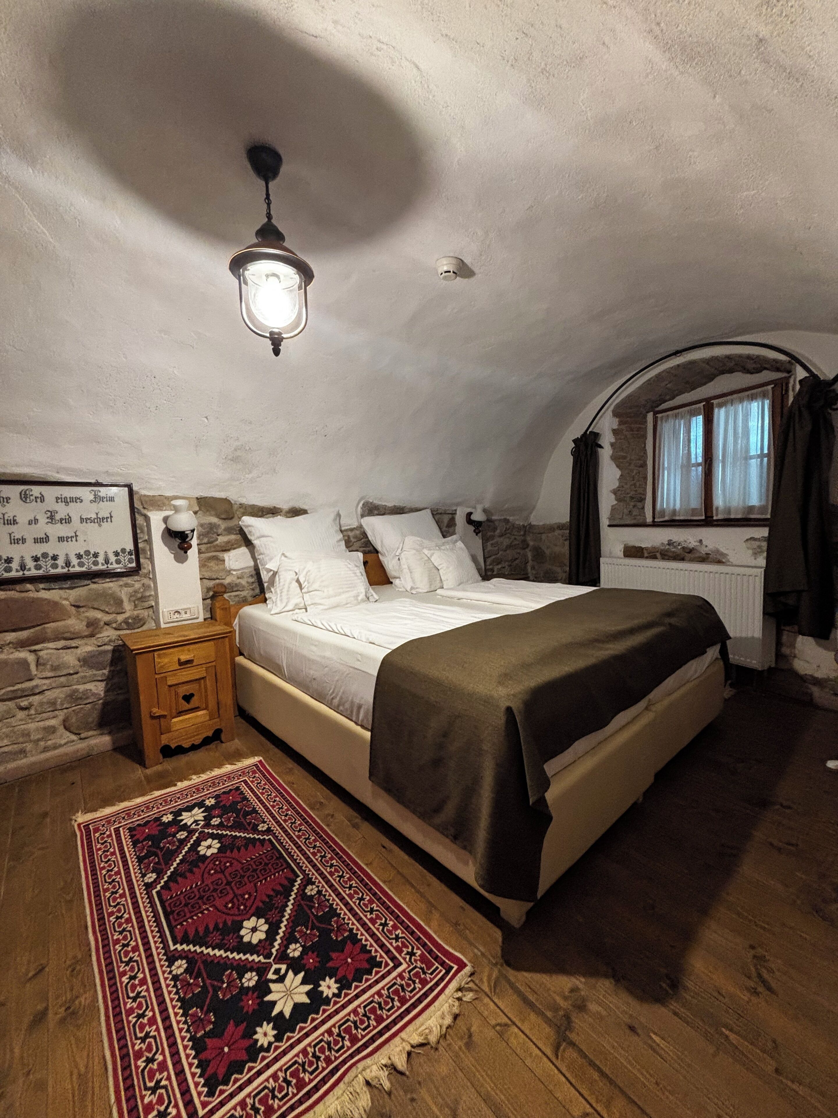 Quarto com cama, mesa de cabeceira, luminária na parede, janela com cortinas e tapete decorativo.
