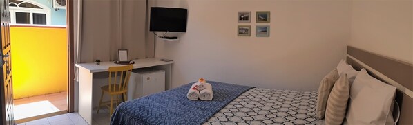 Quarto de hotel com cama, escrivaninha, cadeira, televisão e pequeno frigorífico.