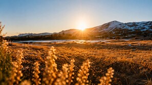 Paisagem montanhosa com nascer ou pôr do sol, um campo de grama e um corpo d'água.