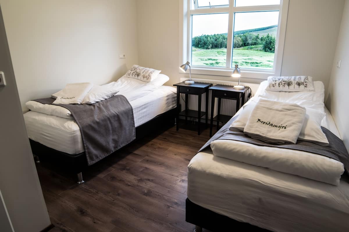 Quarto com duas camas, uma mesa de cabeceira, uma luminária e uma janela com vista para o verde.