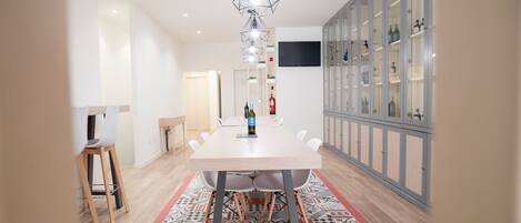 Uma área de jantar moderna com uma mesa comprida, cadeiras, um tapete estampado, um lustre e um armário para vinhos.
