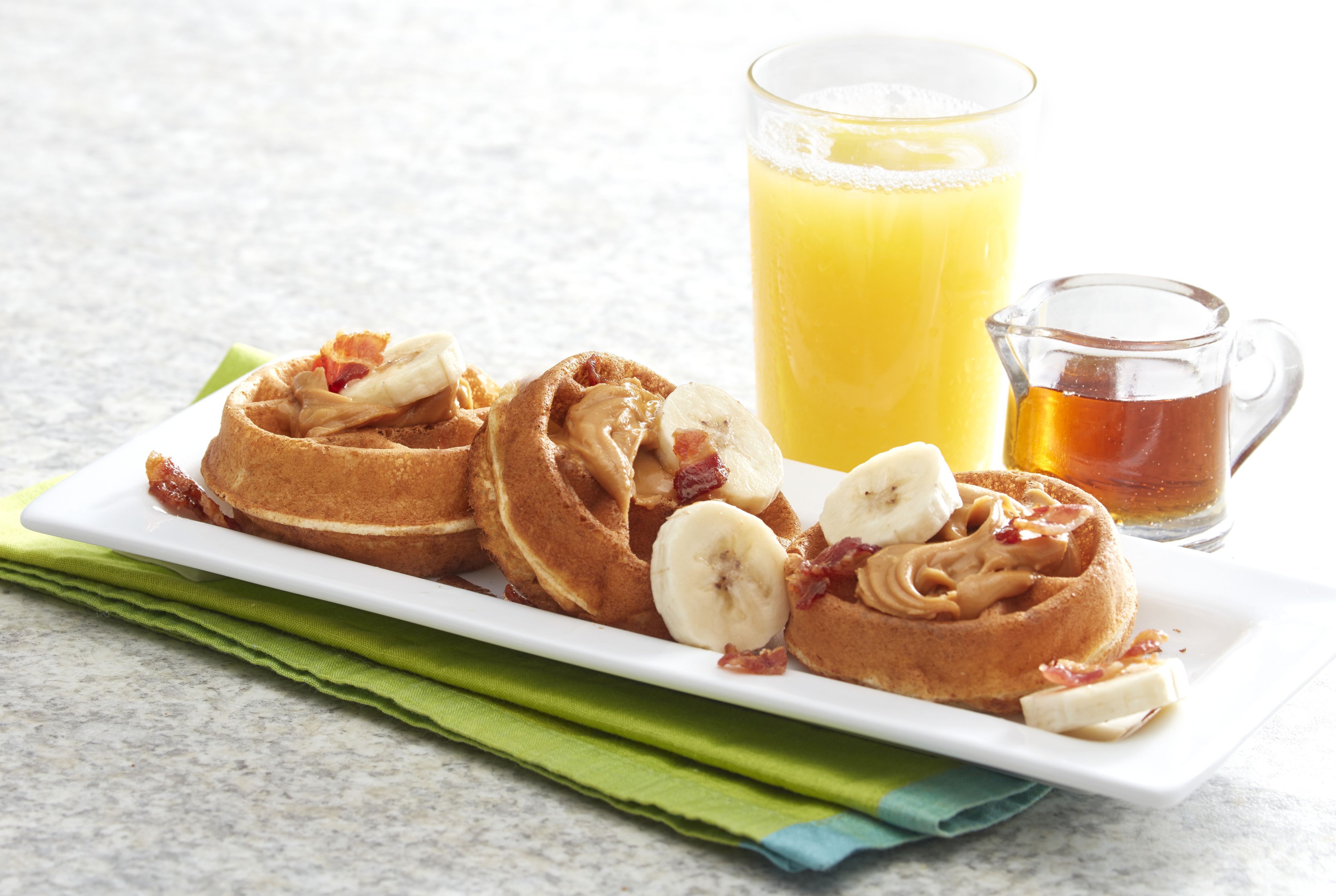 Prato de café da manhã com panquecas, bananas e bacon, acompanhado de um copo de suco de laranja e um jarro de mel.