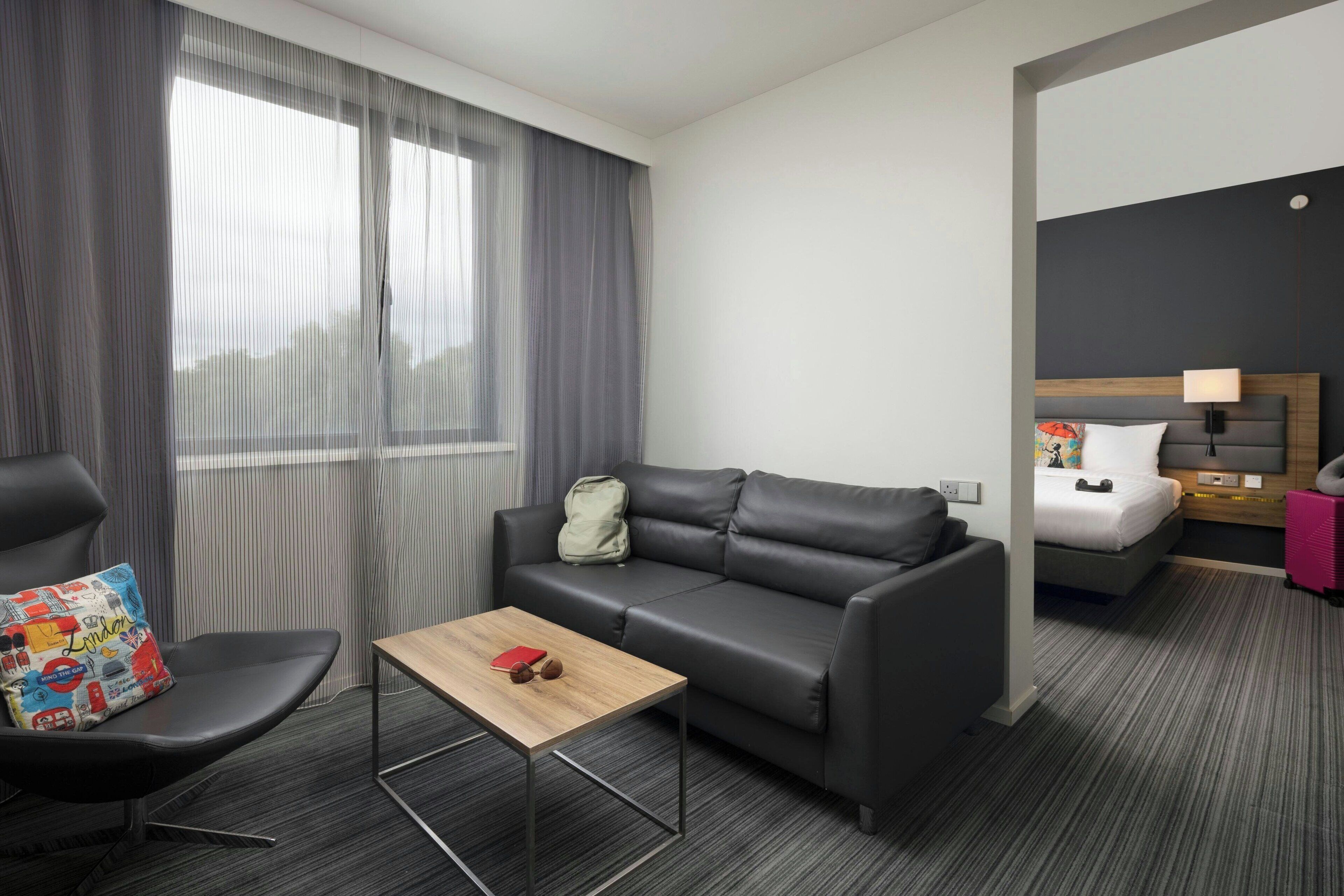 Quarto de hotel moderno com sofá de couro, mesa de centro de madeira, poltrona preta e cama com encosto.