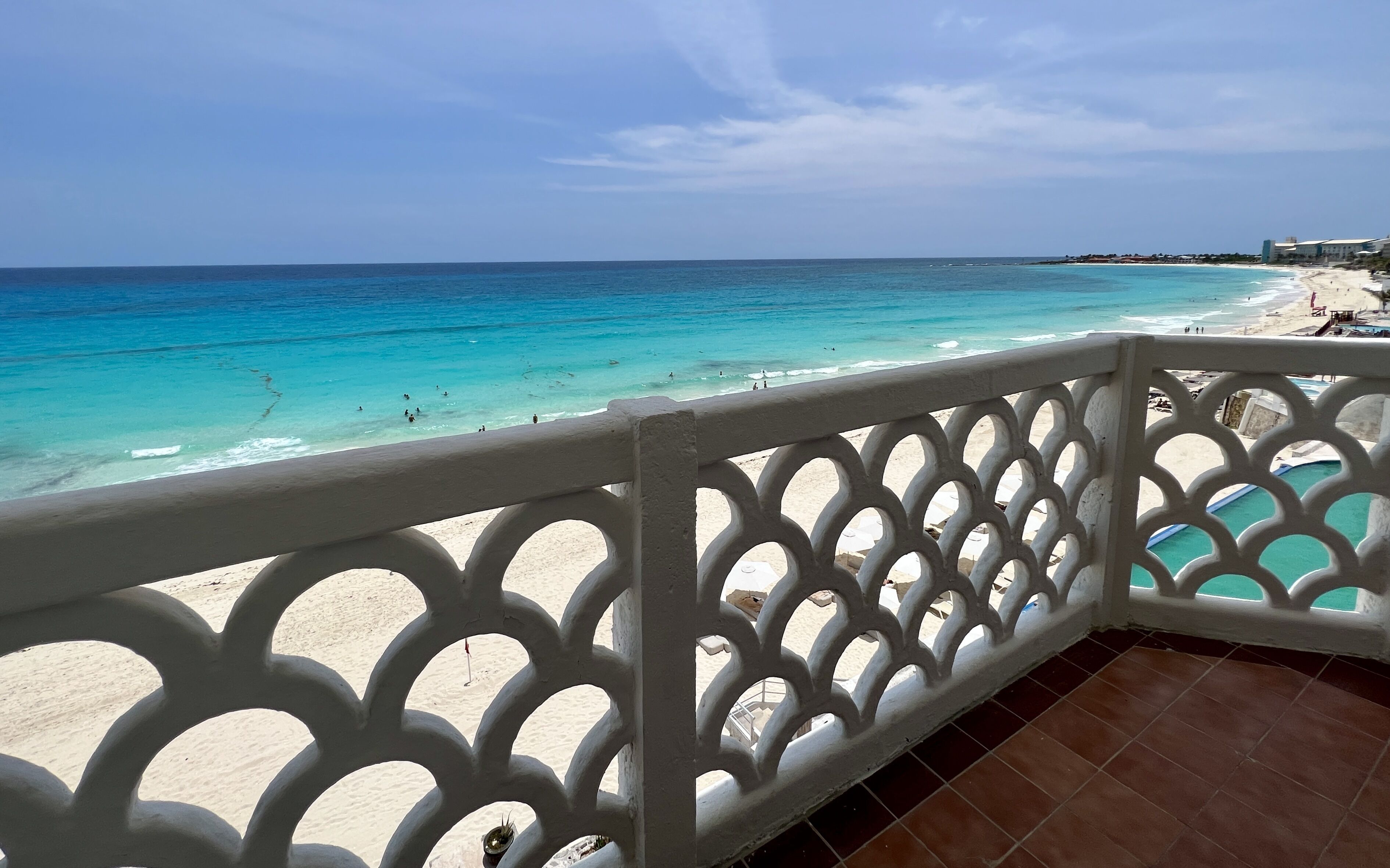 Vista da praia a partir de uma varanda com guarda-corpo branco, água turquesa translúcida e uma praia arenosa.