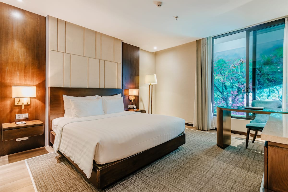 Quarto de hotel moderno com uma cama grande, abajures, uma escrivaninha e vista para árvores pela janela.