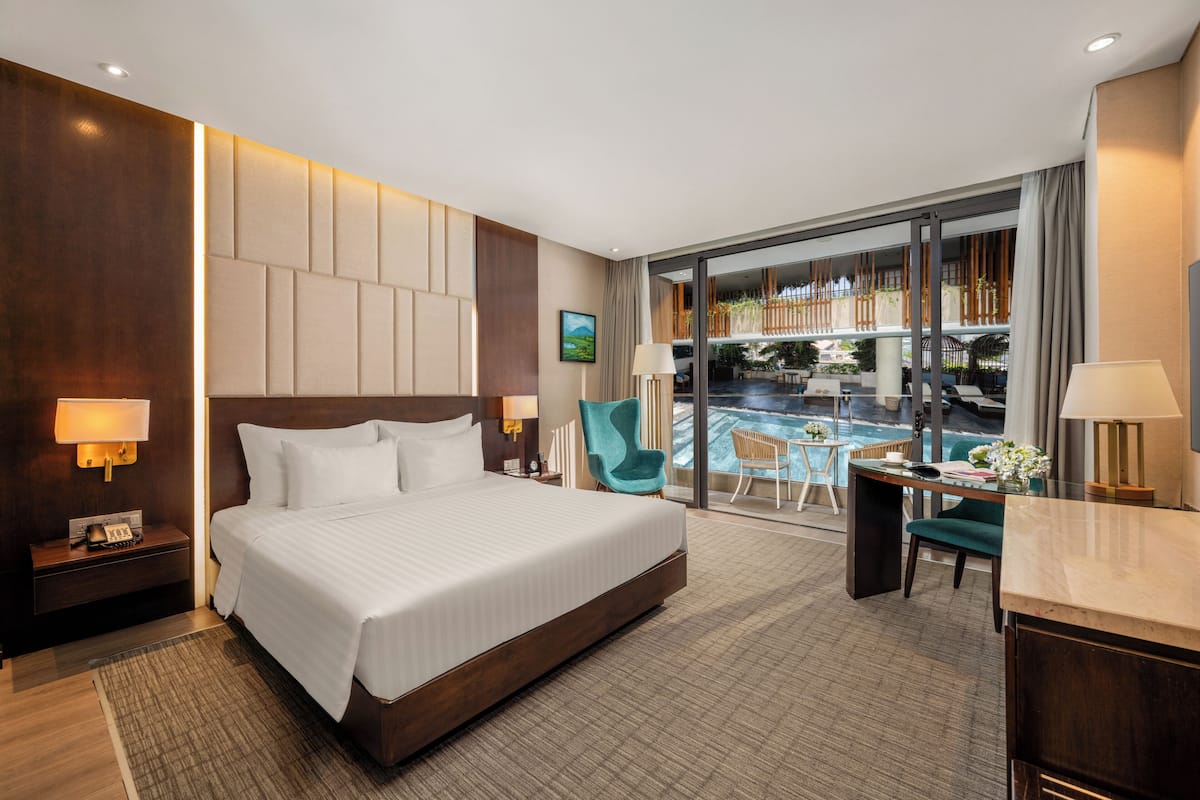 Quarto de hotel com uma cama grande, uma escrivaninha com cadeira, uma mesinha de cabeceira com abajur e vista para a área da piscina.
