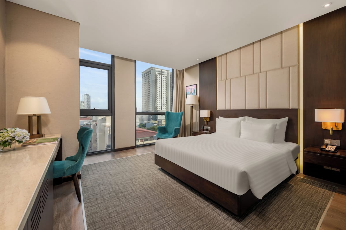 Quarto de hotel com uma cama grande, uma escrivaninha com abajur, uma cadeira e vista para edifícios altos através de uma janela ampla.