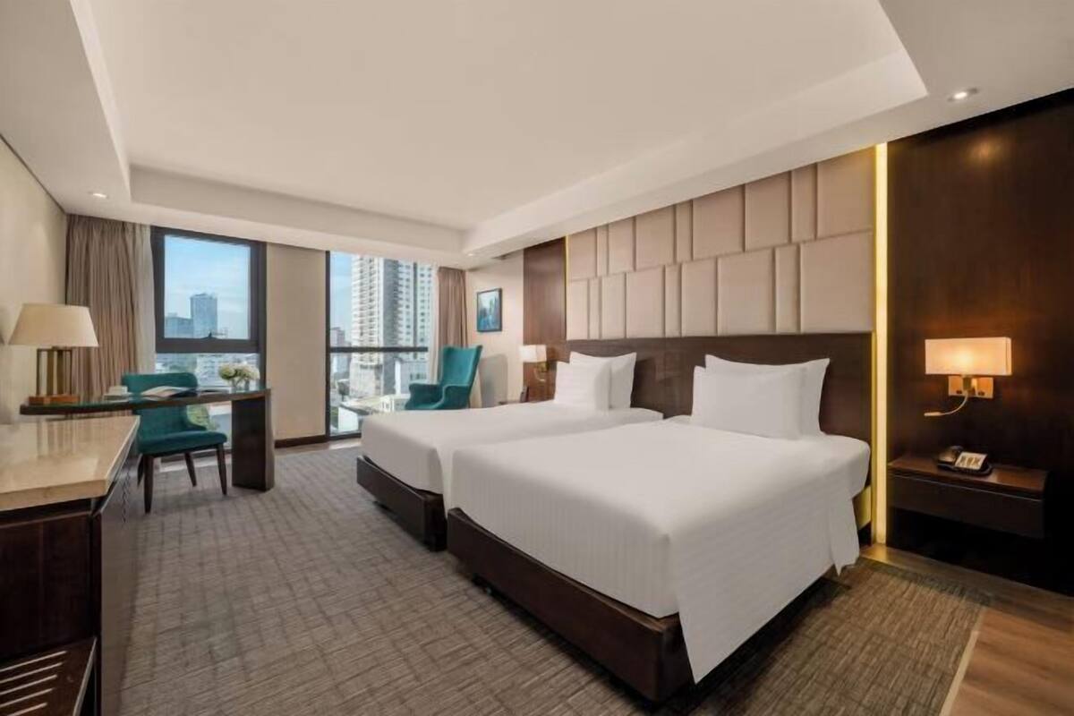 Quarto de hotel com duas camas, uma escrivaninha, uma cadeira, um abajur e vista para a cidade.