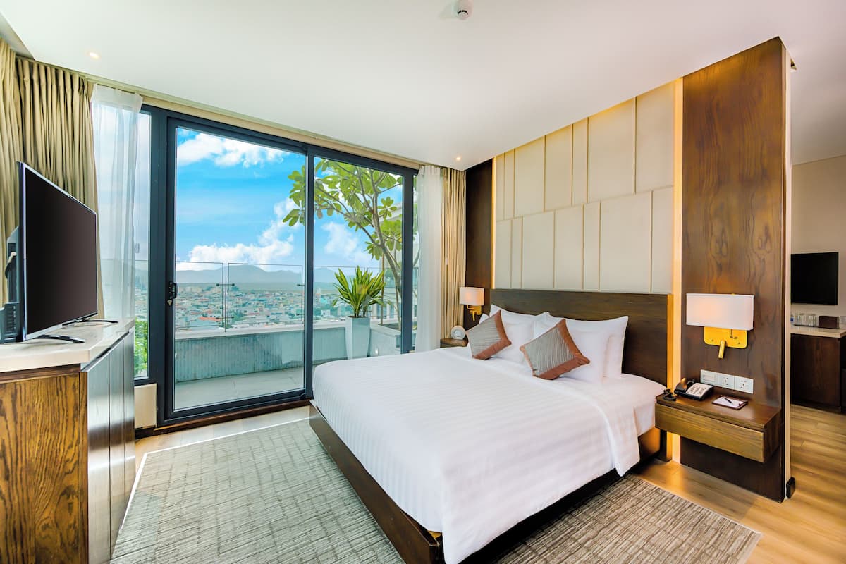 Quarto de hotel moderno com uma cama grande, televisão, escrivaninha e varanda com vista para a cidade.