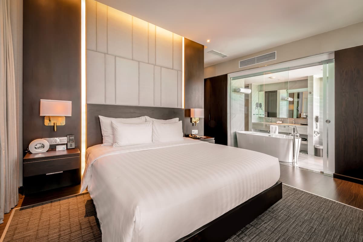 Quarto de hotel moderno com uma cama grande, mesas de cabeceira com luminárias, uma escrivaninha embutida e um banheiro visível por uma porta aberta.
