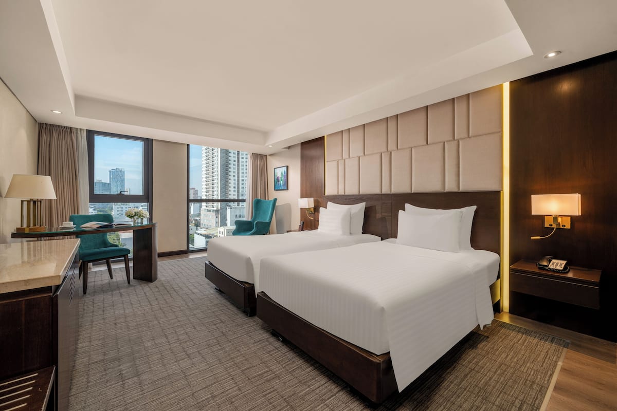 Quarto de hotel com duas camas, uma escrivaninha, uma cadeira e uma janela grande com vista para a cidade.