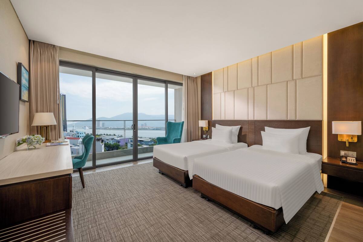 Quarto de hotel com duas camas, uma escrivaninha, uma cadeira, televisão e uma janela grande com vista para a paisagem urbana e para a água.