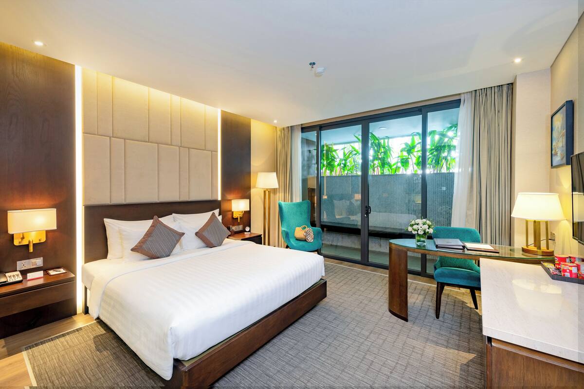 Quarto de hotel com cama grande, escrivaninha, cadeira e vista para o verde pela janela.