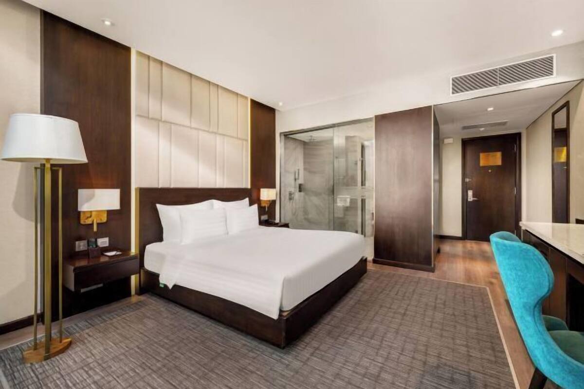 Quarto de hotel com uma cama grande, escrivaninha, cadeira, luminária e box de vidro para o chuveiro.