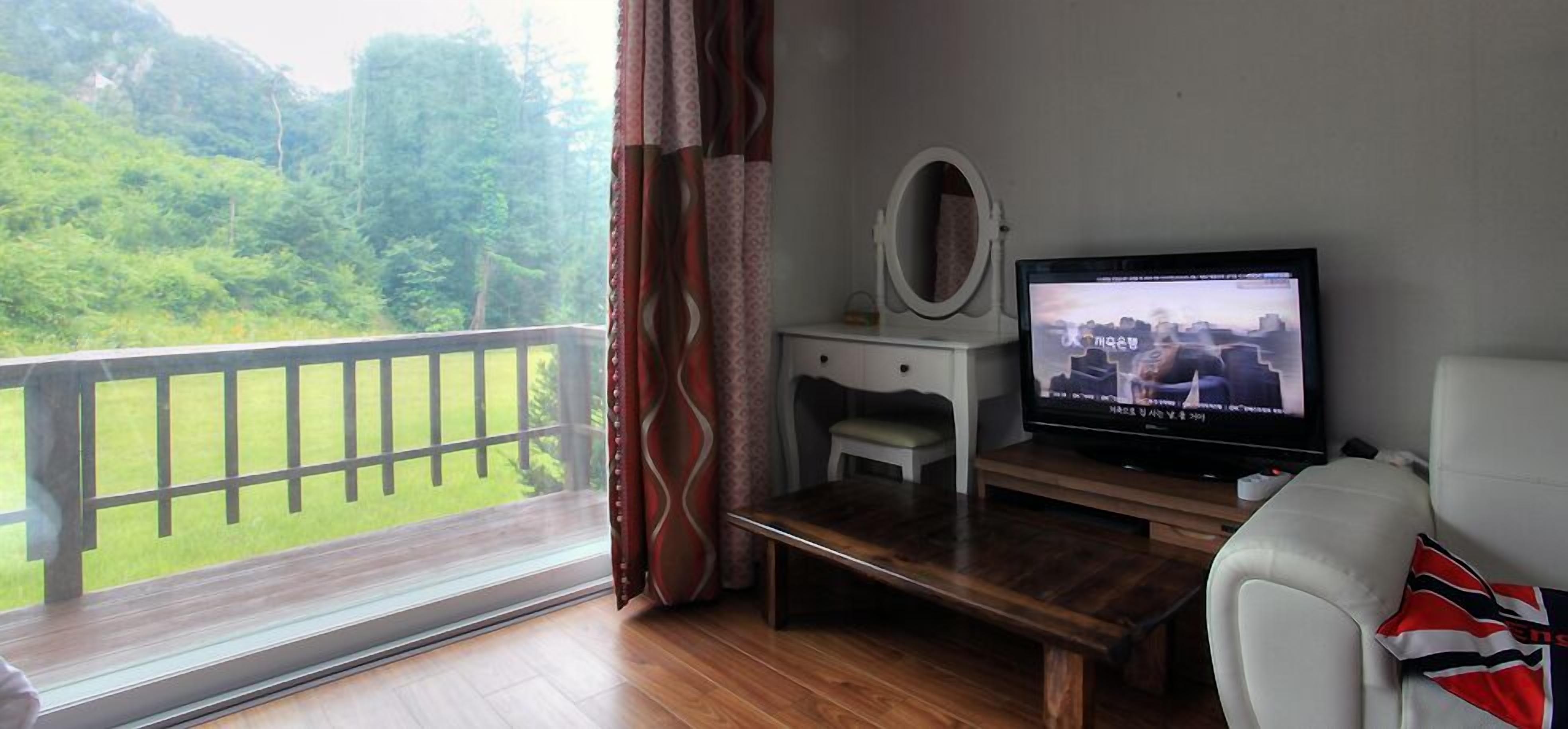 Quarto com piso de madeira, televisão em um suporte, uma mesinha, uma cadeira branca, um espelho e vista para uma varanda com guarda-corpo de madeira e vegetação lá fora.