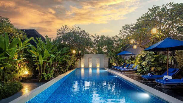 Piscina com espreguiçadeiras e guarda-sóis, cercada por vegetação exuberante e um céu ao pôr do sol.