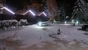 Área externa coberta de neve com mesas de piquenique e bancos, um edifício com uma estrela na fachada e um carro estacionado perto.