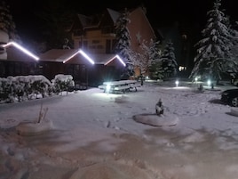 Área externa coberta de neve com mesas de piquenique e bancos, um edifício com uma estrela na fachada e um carro estacionado perto.