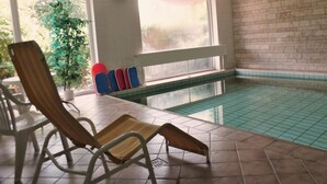 Uma piscina interior com chão de azulejos, um banco de madeira e uma cadeira. Há janelas com vista para o verde lá fora.