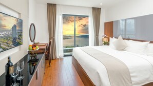 Quarto de hotel moderno com uma cama grande, uma escrivaninha com espelho, vista para o pôr do sol e uma pintura de paisagem urbana.