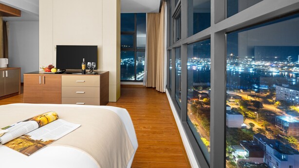 Quarto de hotel moderno com uma cama grande, uma televisão sobre um armário de madeira e vista para a cidade por grandes janelas.