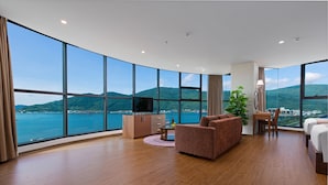 Quarto espaçoso com amplas janelas proporcionando vista para a água e colinas. Há um sofá, uma mesinha e uma televisão.
