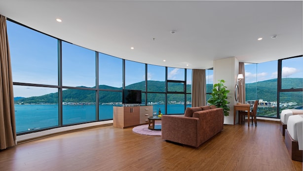 Quarto espaçoso com amplas janelas proporcionando vista para a água e colinas. Há um sofá, uma mesinha e uma televisão.