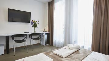 Quarto de hotel com cama, duas cadeiras, escrivaninha, televisão e janela com cortinas.