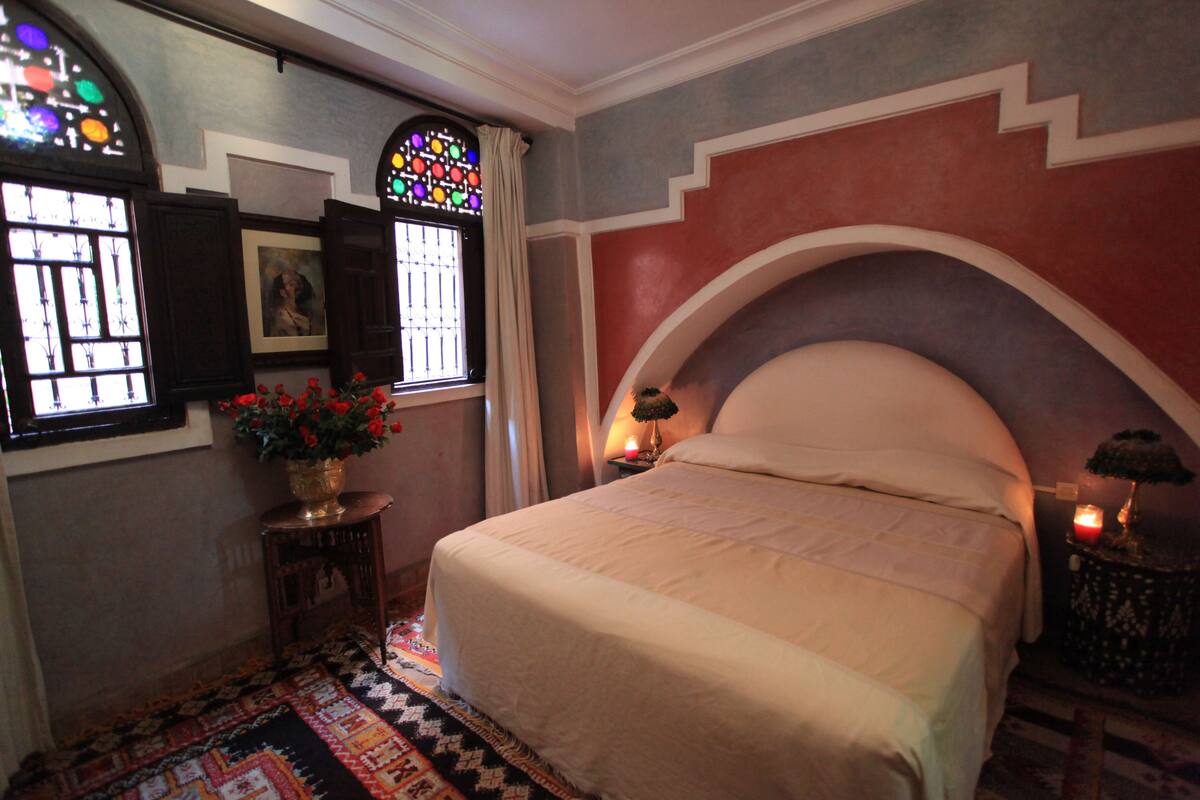 Um quarto com uma cama grande, uma mesinha de cabeceira com abajur, uma pequena mesa com um vaso de flores e uma janela com vitrais.