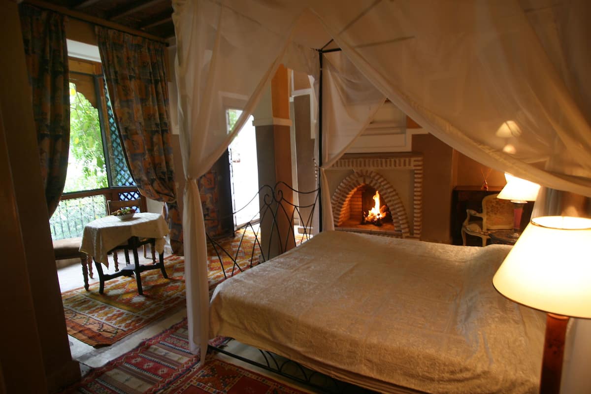Quarto com cama de dossel, lareira, mesa de jantar com cadeiras e vista para o verde lá fora.