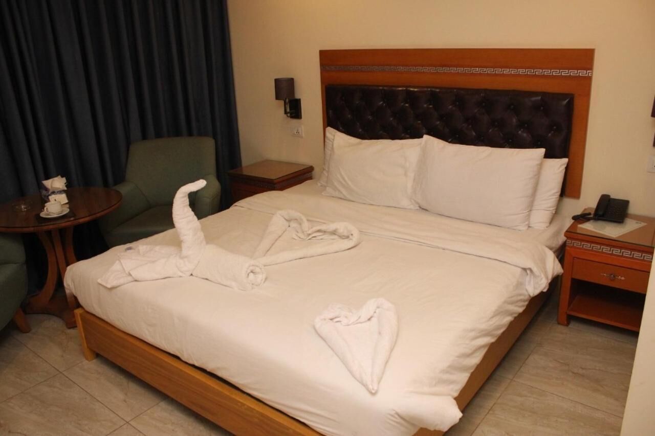 Quarto de hotel com uma cama, uma poltrona verde, uma mesinha com uma chávena e um lenço, e uma cómoda com uma lâmpada.
