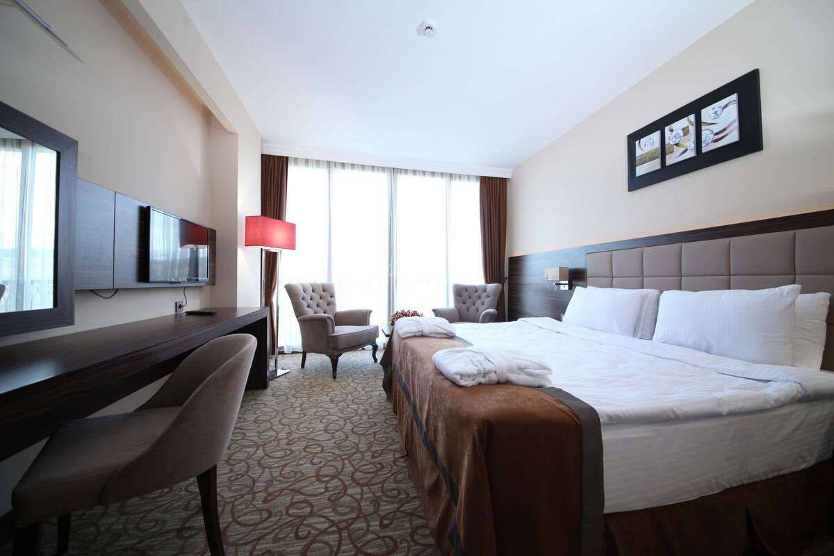 Quarto de hotel com cama, escrivaninha, cadeiras, televisão e janela com cortinas.